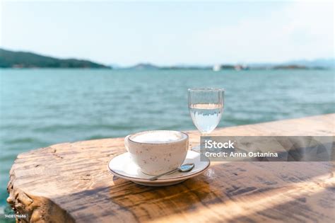 카푸치노 라떼 커피 컵 머그잔과 물 한 잔이 바깥 카페의 나무 테이블에 서서 바다와 산을 배경으로 풍경을 감상합니다 빈 공간 복사 우유에 대한 스톡 사진 및 기타 이미지