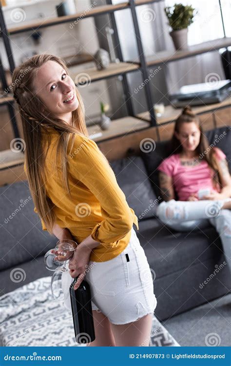 Smiling Lesbian Holding Bottle Of Wine And Wine Glasses While Standing Near Girlfriend In Living