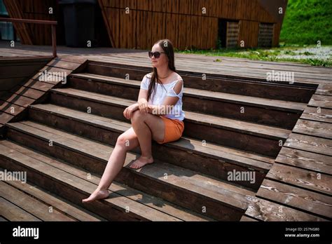 A Barefoot Russian Woman In Her 40s Sits On Wooden Stairs In Summer Wearing Orange Shorts And A