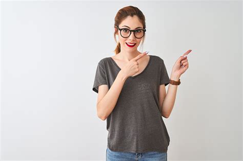 캐주얼 한 티셔츠와 안경을 쓰고 고립된 흰색 배경 위에 서서 미소를 지으며 두 손과 손가락으로 측면을 가리키는 카메라를 바라보고 있는 빨간 머리 여성 측면 보기에 대한 스톡