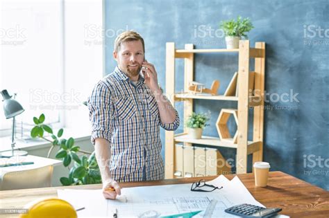 블루 프린트와 카메라를 보면서 휴대 전화에서 고객에 게 얘기 하는 수염과 쾌활 한 흥분 젊은 엔지니어 건설 산업에 대한 스톡 사진 및 기타 이미지 Istock