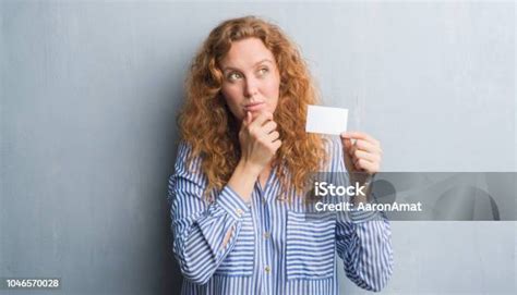 들고 빈 회색 그런 지 벽에 빨강 머리 젊은 여 자가 방문 카드 심각한 얼굴 생각 질문 매우 혼란 스 러 워 아이디어에 대 한 개념에 대한 스톡 사진 및 기타 이미지