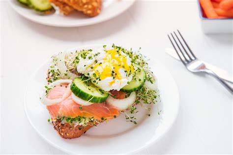 달걀 베네딕트 훈제 연어와 시리얼 롤빵 마이크로 녹색 양파와 오이 조각 크림 치즈 제공된 흰색 나무 테이블에 새싹 건강 한 아침 식사 개념입니다 선택적 초점입니다 갈색 빵에