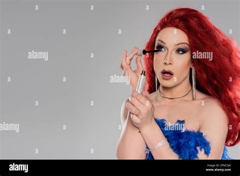 Drag Queen In Red Wig And Dress Applying Mascara Isolated On Greystock