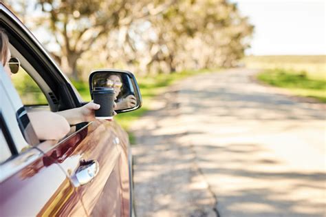 창문 밖에서 한쪽 팔을 들고 검은 커피 컵을 들고 차 안에 앉아있는 여성 종이 커피 잔을 운전하고 들고있는 여성 나무를 지나가는 길에서의 여행 거울에 대한 스톡 사진 및