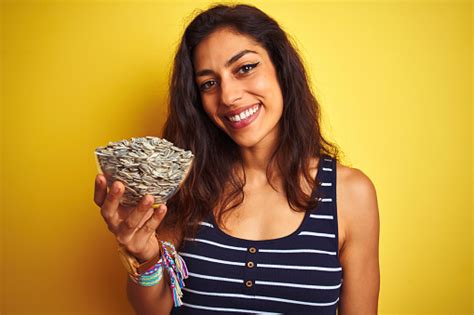 행복한 얼굴 서서 치아를 보여주는 자신감 미소와 함께 미소와 고립 된 노란색 배경 위에 해바라기 씨앗그릇을 들고 젊은 아름다운 여자 갈색 머리에 대한 스톡 사진 및 기타