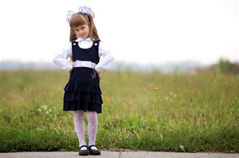 흐리게 밝은 녹색 햇볕이 잘 드는 잔디와 흰색 하늘 복사 공간 배경에 긴 금발 머리에 학교 유니폼과 흰색 활귀여운 사랑스러운 심각한 사려 깊은 1 학년 소녀의 전체 길이