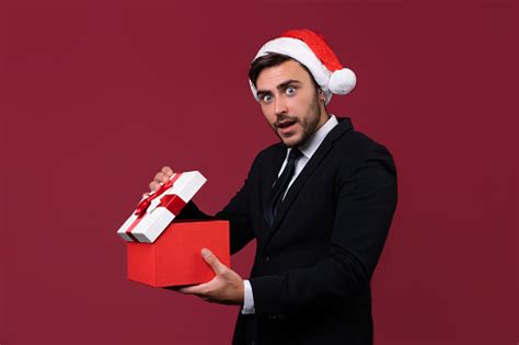 비즈니스 슈트와 산타 모자를 입은 젊은 잘생긴 백인 남자는 스튜디오와 치아가 빨간 선물 상자를 손에 들고 빨간 배경에 서 있습니다 남자 사업가에 대한 스톡 사진 및 기타