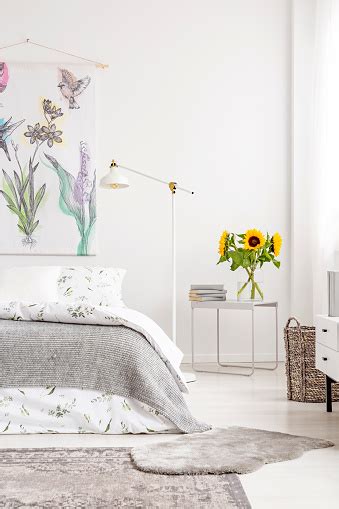 회색 양탄자와 카펫 램프와 재료 회화와 흰색 침실 인테리어의 실제 사진에 꽃 시트와 침대에 서 있는 유리 화병과 책에 신선한 해바라기와 금속 머리 맡 테이블 가구에 대한