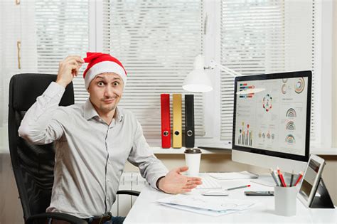 잘생긴 충격과 난처 한 비즈니스 문서 창 배경에 빛 사무실에 현대 모니터 컴퓨터에서 작동과 셔츠와 레드 크리스마스 모자는 책상에 앉아 있는 남자 새 해입니다 경영자에 대한