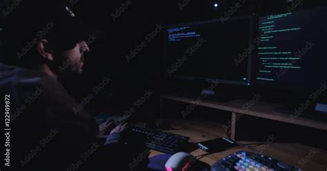 Cyber Terror Computer Hacker Sitting In A Dark Room In Front Of Screens And Writing Lines Of