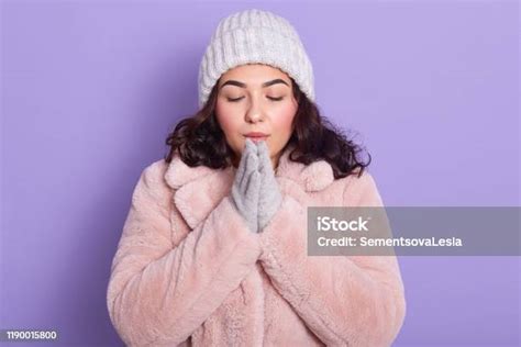 겨울에 손을 잡고 따뜻한 옷 인조 모피 코트 장갑 모자를 쓰고 라일락 배경으로 고립 된 포즈를 취한 아름다운 젊은 여성이 눈을 감고 서 있습니다 겨울과 추운 날씨 개념입니다