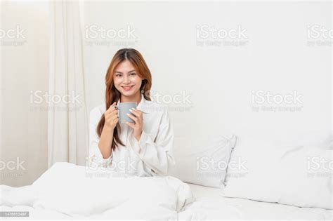 하얀 잠옷을 입은 여자가 주말 아침에 일어나 휴식을 취하고 노트북 휴대폰을 가지고 놀며 하얀 침실 안의 유리에서 빵을 먹고 차를 마신다 아침 휴가 개념입니다 갈색 머리에