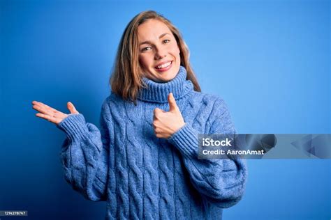 젊은 아름다운 금발의 여자 를 입고 캐주얼 터틀넥 스웨터 위에 푸른 배경 손바닥을 보여주는 엄지 손가락으로 확인 제스처를하고 행복하고 쾌활한 미소 겨울에 대한 스톡 사진 및