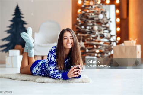 축 하 및 휴일 개념입니다 매력적인 귀여운 여자 니트 휴가 스웨터에 장식 된 크리스마스 트리 근처 계 피와 함께 따뜻한 차 한잔과 함께 흰색 카펫에 거짓말 가정의 방에 대한