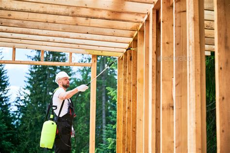 Worker Applying Fire Retardant Using Sprayer While Constructing Wooden Frame House Near Forest