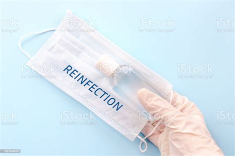 보호 장갑을 낀 손에는 파란색 배경에 Reinfection 및 방부제라는 단어가 있는 의료 용 마스크를 들고 있습니다 코로나바이러스 코피드19를 가진 가능한 재감염의 개념