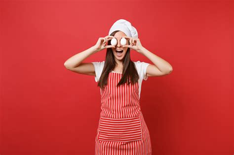 주부 여성 요리사 요리사 또는 줄무늬 앞치마 흰색 티셔츠 모자 요리사 모자 빨간 벽 배경에 고립 된 베이커 재미 있는 정부 여자 잡고 손에 두 계란 복사 공간 개념을 비웃는