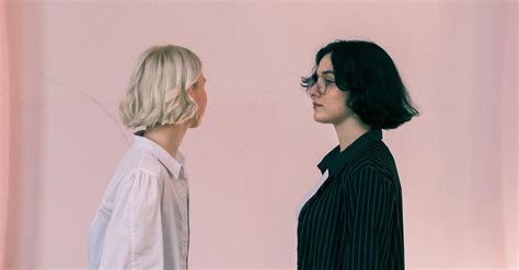 가판대 검은 머리 고요한 금발 대조 두 사람 무표정의 미니멀리스트 미묘한 색상 부드러운 톤 분홍색 배경 색상 조정 서로 마주 세련된 복장 소프트