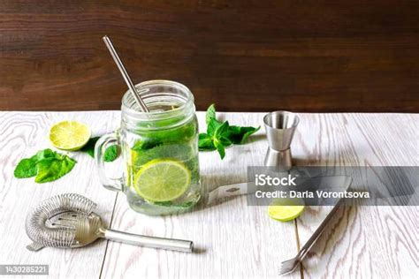 신선한 아이스 콜드 모히토 레모네이드 여름 음료 얇게 썬 라임 칵테일 투명한 컵에 녹색 민트 잎 나무 테이블에 메이슨 항아리 셰이커 지거 스트레이너 숟가락 바만 액세서리