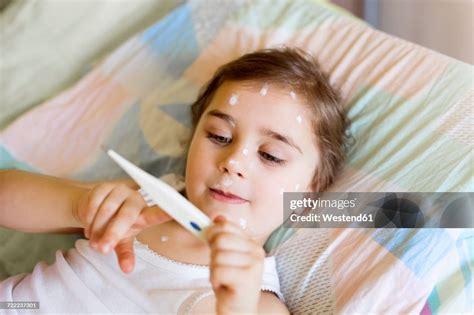 Girl Having Chickenpox Lying In Bed Looking At Clinical Thermometer