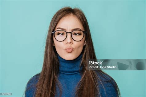 젊은 금발 이탈리아 여성 제스처 입과 쪼그리고 앉는 눈 미친 얼굴 청록색 배경에 대한 코믹한 분위기로 물고기 얼굴을 만드는 제스처 스튜디오에서 파란색 스웨터와 안경을 쓴