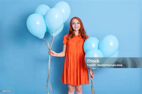 풍선 우아한 드레스를 입고 빨간 물결 모양의 머리를 가진 예쁜 여자는 카메라를 보고 그녀의 생일을 축하하는 행복한 여자 파란색 배경 위에 고립 되어 서 20 24세에 대한