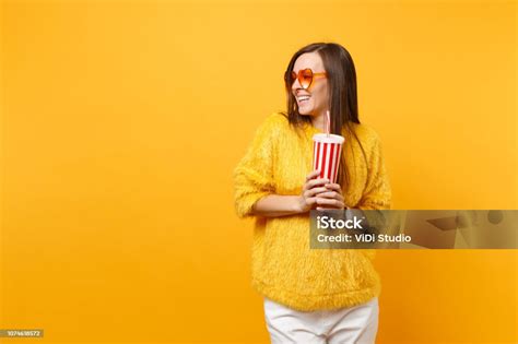 모피 스웨터에서 즐거운 젊은 여자 심장 오렌지 안경 보고 옆으로 콜라 나 소 다 지주 플라스틱 컵 고립 노란색 배경에 사람들이 진심으 감정 라이프 스타일 개념 광고