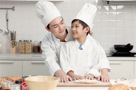 주방에서 모자를 쓰고 요리사 유니폼을 입은 행복한 아시아인 아들과 아버지 소년은 밀대로 반죽을 반죽하고 아빠는 아이가 빵을 굽는 것을 돕고 함께 빵집을 만드는 동안 서로를