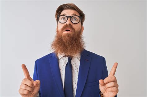 고립 된 흰색 배경 위에 양복과 안경을 착용한 젊은 빨간 머리 아일랜드 사업가는 손가락과 팔을 들고 위를 올려다 보고 놀랐습니다 가리키기에 대한 스톡 사진 및 기타 이미지