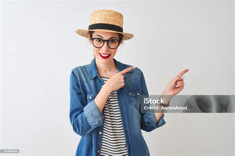 데님 셔츠 안경과 모자를 쓰고 고립된 흰색 배경위에 미소를 지으며 두 손과 손가락으로 측면을 가리키는 카메라를 바라보는 빨간 머리 여성 가리키기에 대한 스톡 사진 및 기타