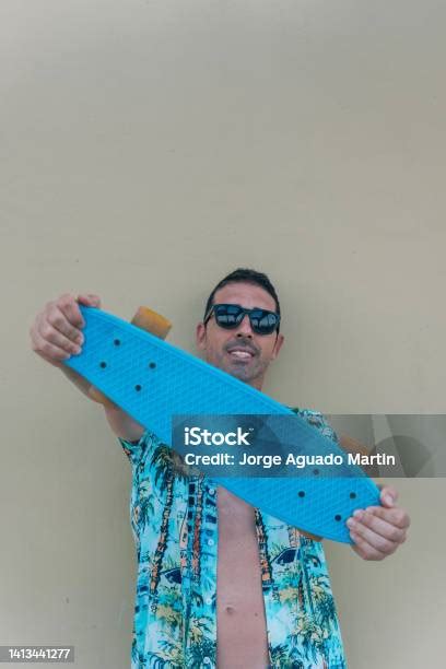 밝은 색의 벽에 기대어 파란 셔츠와 검은 색 선글라스를 착용하고 푸른 페니 롱 보드를 보여주는 젊은 웃는 갈색 머리 남자의 수직