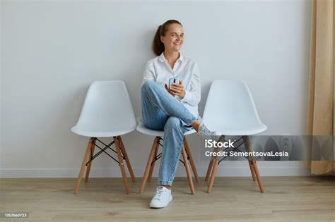 이빨이 많은 미소를 지으며 행복한 여자 손에 스마트폰을 들고 카메라를 바라보며 의자에 앉아 청바지와 흰색 셔츠를 입고 긍정적인 감정을 표현하고 사람들에 대한 스톡 사진 및