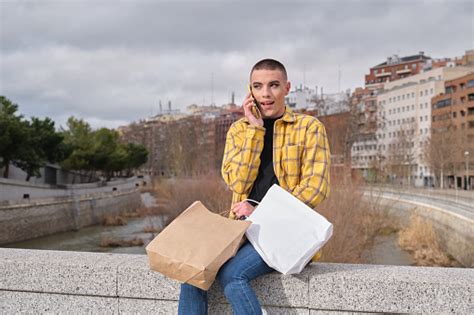 잘생긴 젊은 남자는 화장을 입고 미소와 쇼핑 백을 들고 자신의 스마트 폰에 말하기 놀랐다 비 바이너리 앤드로기누스 사람 20 29세에 대한 스톡 사진 및 기타 이미지