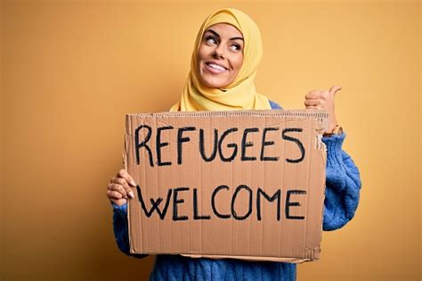 아름 다운 아랍 여자 를 입고 이슬람 히잡 들고 배너 와 난민 환영 메시지 가리키는 하 고 행복 한 얼굴 미소와 함께 측면에 엄지 손가락으로 표시 가리키기에 대한 스톡 사진