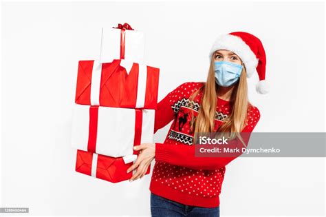 크리스마스 산타 클로스 모자를 쓰고 얼굴에 의료 용 마스크를 쓰고 크리스마스 선물과 함께 흰색 배경에 산타의 도우미를 입은 젊은 행복한 여성 12월에 대한 스톡 사진 및