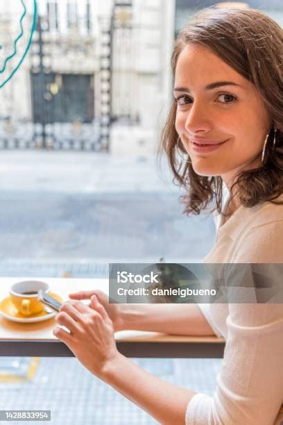 창문 옆 바에 앉아 스마트폰을 손에 들고 테이블에 커피 한 잔을 들고 카메라를 향해 웃고 있는 젊은 갈색 머리 여인 카페에 대한