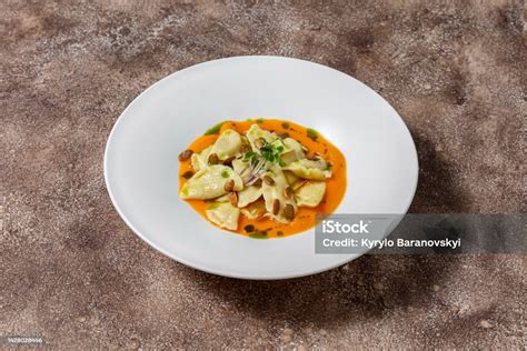 포르치니 만두 호박 소스 호박씨 발사믹 페스토 소스 마이크로그린 콩나물 음식은 흰색 둥글고 깊은 세라믹 접시에 놓여 있습니다 접시는 어두운 패브릭 배경 커피 색상에 서