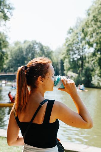 화창한 여름날 공원에서 야외에 서 있는 컵을 마시며 눈을 감고 있는 검게 그을린 젊은 여성 물과 나무 근처의 아름다운 빨간 머리