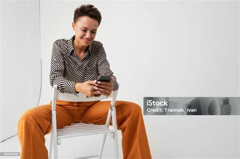 짧은 어두운 머리 검은 색과 흰색 줄무늬 셔츠 갈색 바지와 신발에 메이크업과 젊은 아름다운 소녀는 그녀의 손에 그녀의 휴대 전화와 의자에 앉아 생각 앉음에 대한 스톡 사진