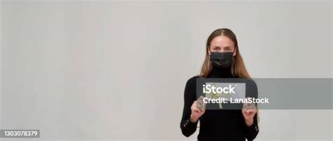 검은 색 의상과 보호 마스크를 착용한 젊은 백인 여성이 회색 배경에 고립 된 포즈를 취하면서 쇼핑을위한 상품권을 들고 카메라를 보고 있습니다 169에 대한 스톡 사진 및