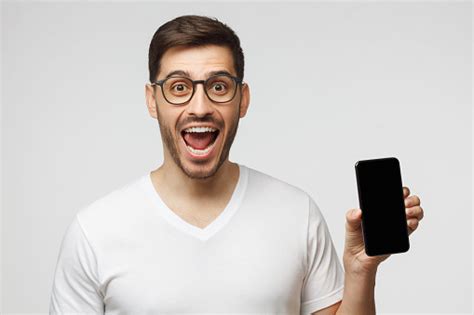 흰색 티셔츠와 안경을 쓴 젊은 흥분한 남자가 회색 배경에 고립된 깜짝 얼굴을 보며 빈 화면 스마트폰을 보는 모습을 볼 수 있습니다 T 셔츠에 대한 스톡 사진 및 기타 이미지