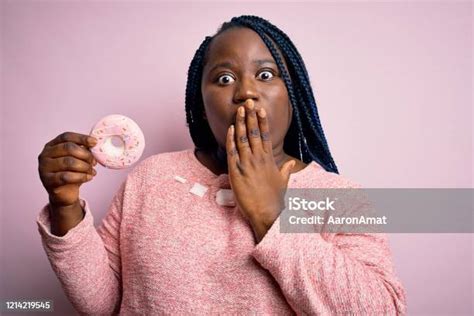 젊은 아프리카 계 미국인 플러스 크기 여자 와 머리띠 먹는 도넛 위에 분홍색 배경 커버 입 손으로 충격 실수 두려움의 표정