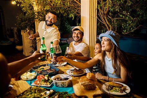 가족과 친구들이 수영장 옆 스트링 조명 아래 야외 테이블에서 식사를하고 있습니다 음식과 음료가 있는 저녁 사교 모임 여름 축하 컨셉 미디엄 샷 25 29세에 대한 스톡 사진