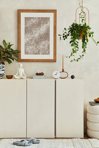 모의 포스터 프레임 베이지 색 나무 사이드 보드 푸프 식물및 보헤미안 에서 영감을 받은 개인 액세서리가있는 창의적이고 현대적인 베이지 색 거실 인테리어 디자인 공간을