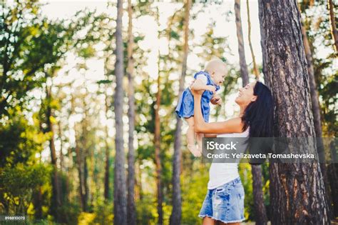 여름 공원에서 침 엽 수 숲에서 파란 눈에 금발 딸의 출생의 1 년을 잡고 긴 갈색 머리를 가진 젊은 아름 다운 세련 된 어머니 가족에 대한 스톡 사진 및 기타 이미지