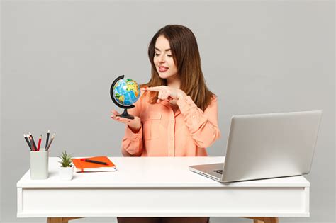 지구 세계 글로브에 인덱스 손가락을 가리키는 젊은 예쁜 여 자가 앉아서 회색 배경에 고립 된 Pc 노트북과 흰색 책상에서 작동 합니다 성취 비즈니스 커리어 라이프 스타일