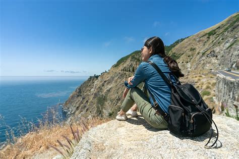 화창한 날 바위에 배낭을 메고 앉아 해안선 하이웨이 근처에서 평화로움을 즐기는 잔잔한 바다를 바라보는 아시아 한국 소녀의 뒷모습 고속도로에 대한 스톡 사진 및 기타 이미지