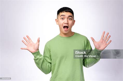 초록색 스웨트셔츠를 입은 흥분한 아시아 남성이 손바닥을 활짝 벌리고 즐거운 표정으로 양손을 들어 올린다 그는 평범한 흰색 배경에 서서 놀라움과 흥분을 전달합니다 30 34