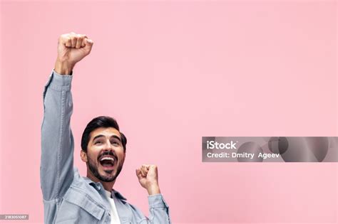 턱수염 행복 승리를 가진 갈색 머리 남자의 패션 초상화는 흰 티셔츠 미소와 그의 얼굴에 기쁨의 감정에 분홍색 배경에 주먹을 들고